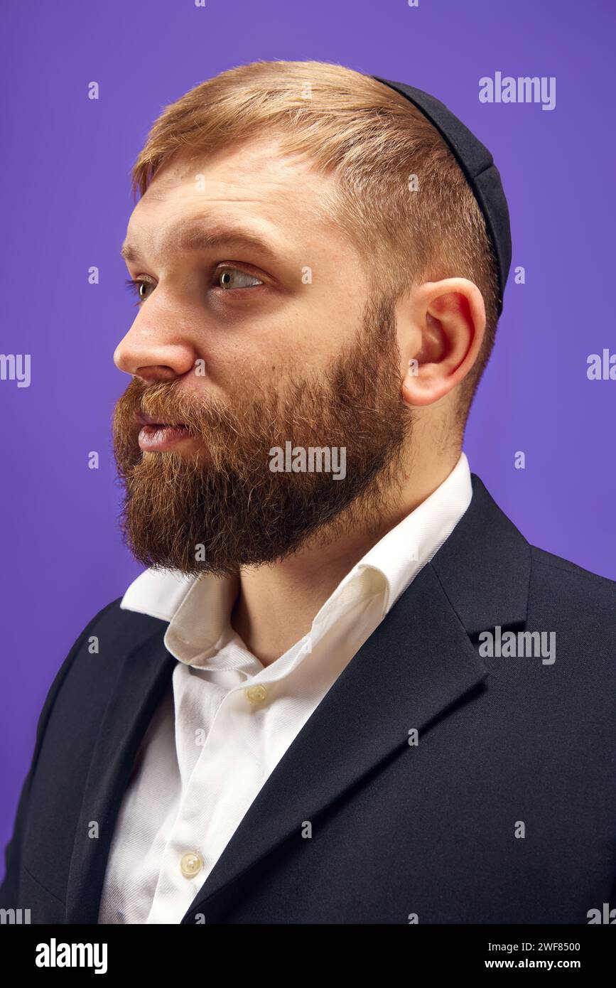 Portrait of bearded young Jewish man in yarmulke posing against purple ...