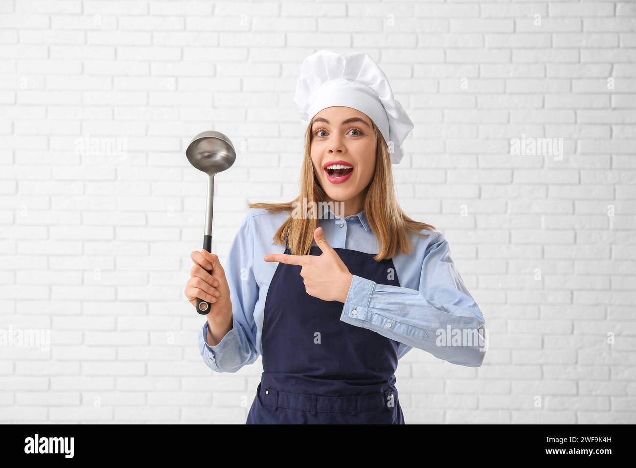 Young female chef pointing at ladle on white brick wall background ...