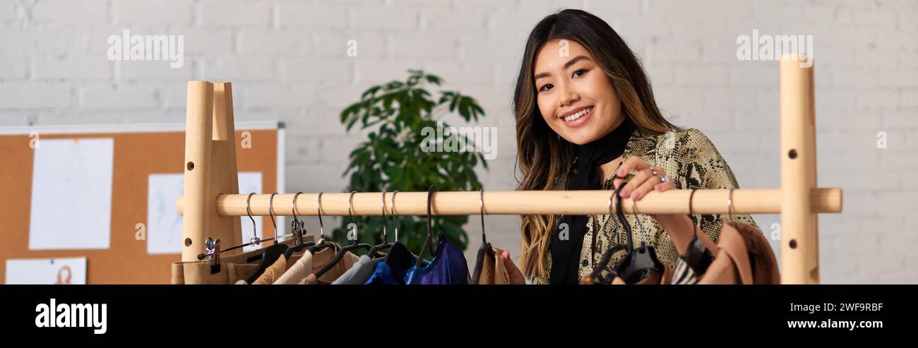 joyful asian dressmaker smiling at camera near rack with bespoke ...