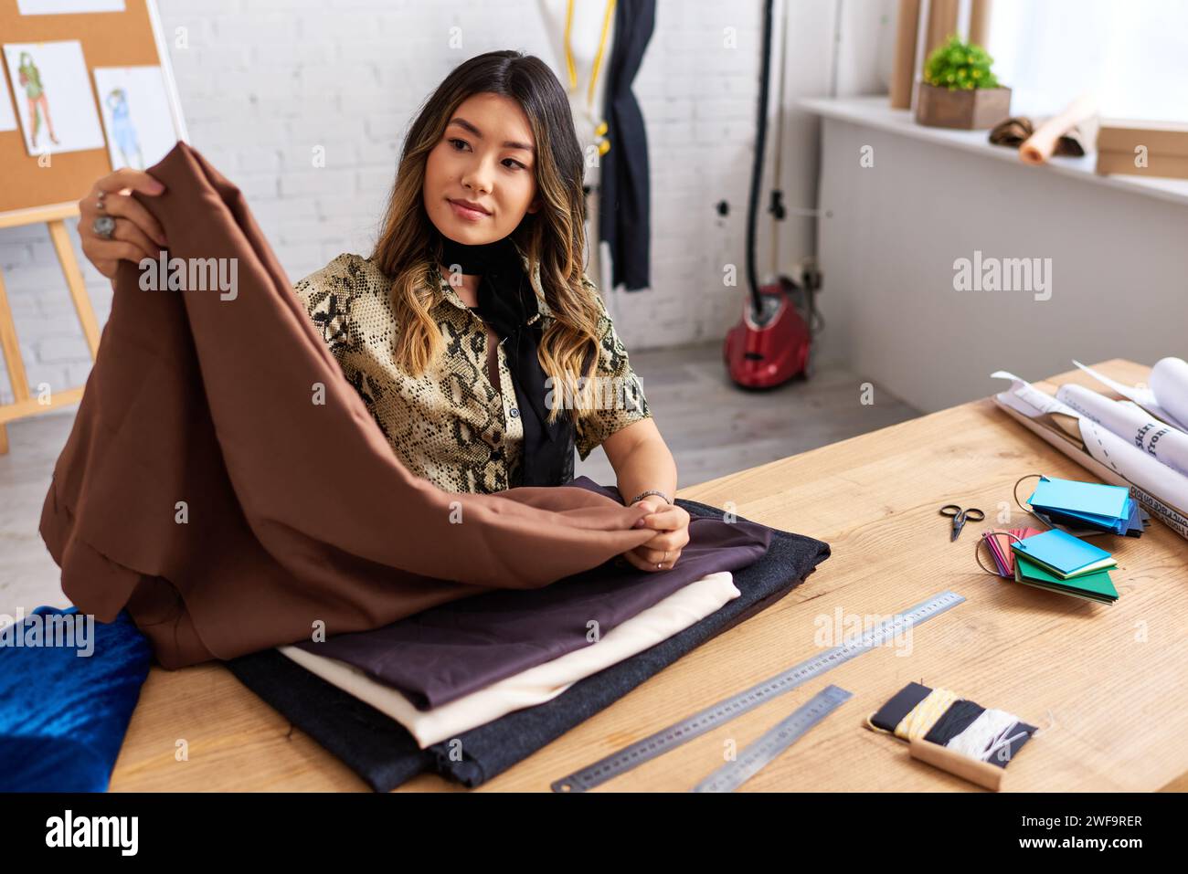 young creative asian stylist looking at fabrics samples in own atelier ...