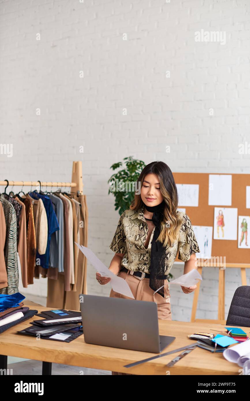 creative asian stylist looking at sewing pattern near laptop and rack ...