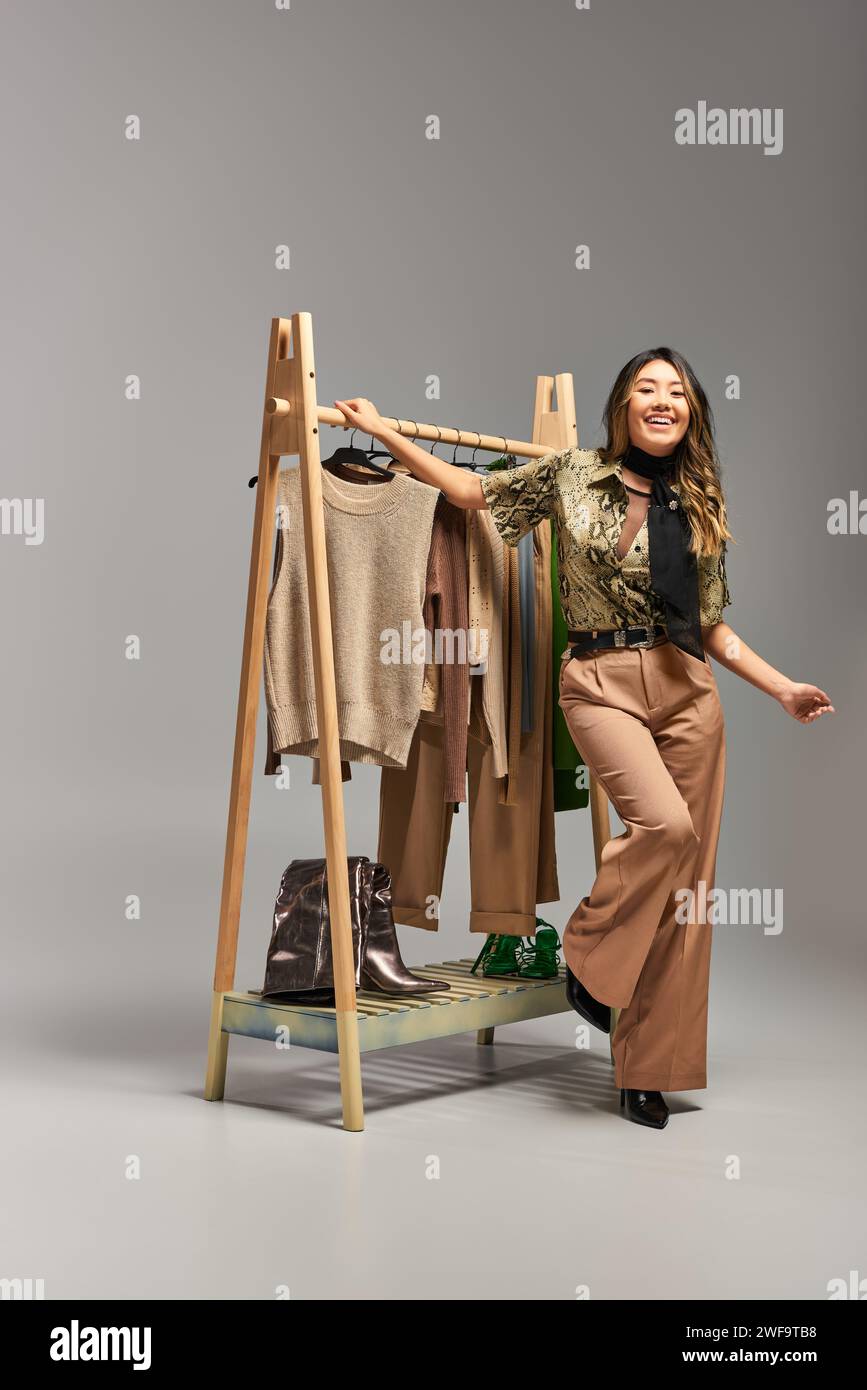 overjoyed asian stylist smiling near rack with bespoke clothes on grey ...