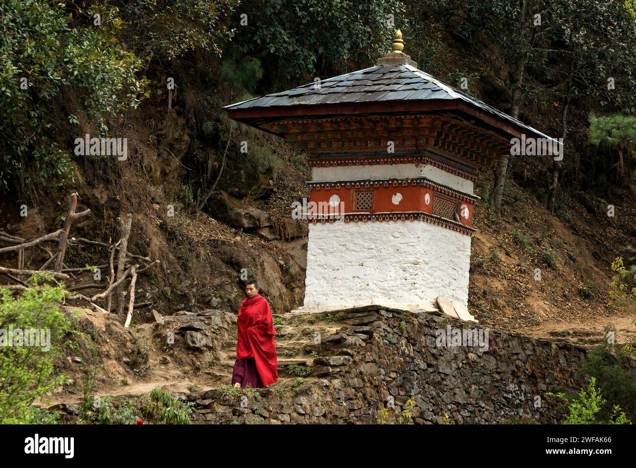 A monk in a red robe descends a rock path past a small chorten as he ...