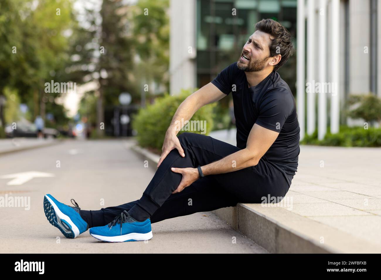 A young sportsman man sits on the street on the curb and holds his leg ...