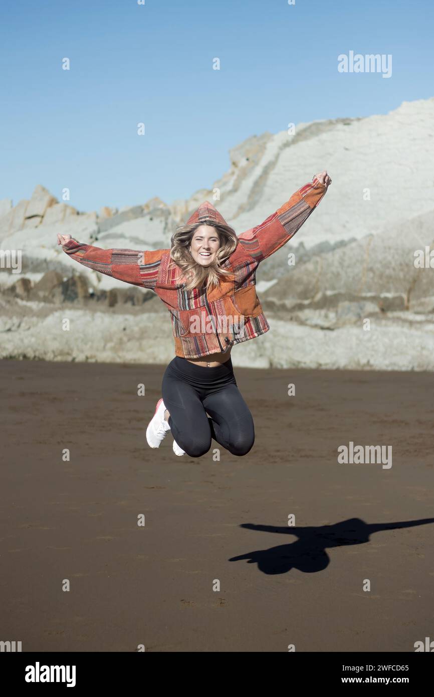 woman is joyfully jumping on a beach, casting a shadow, with rocky ...