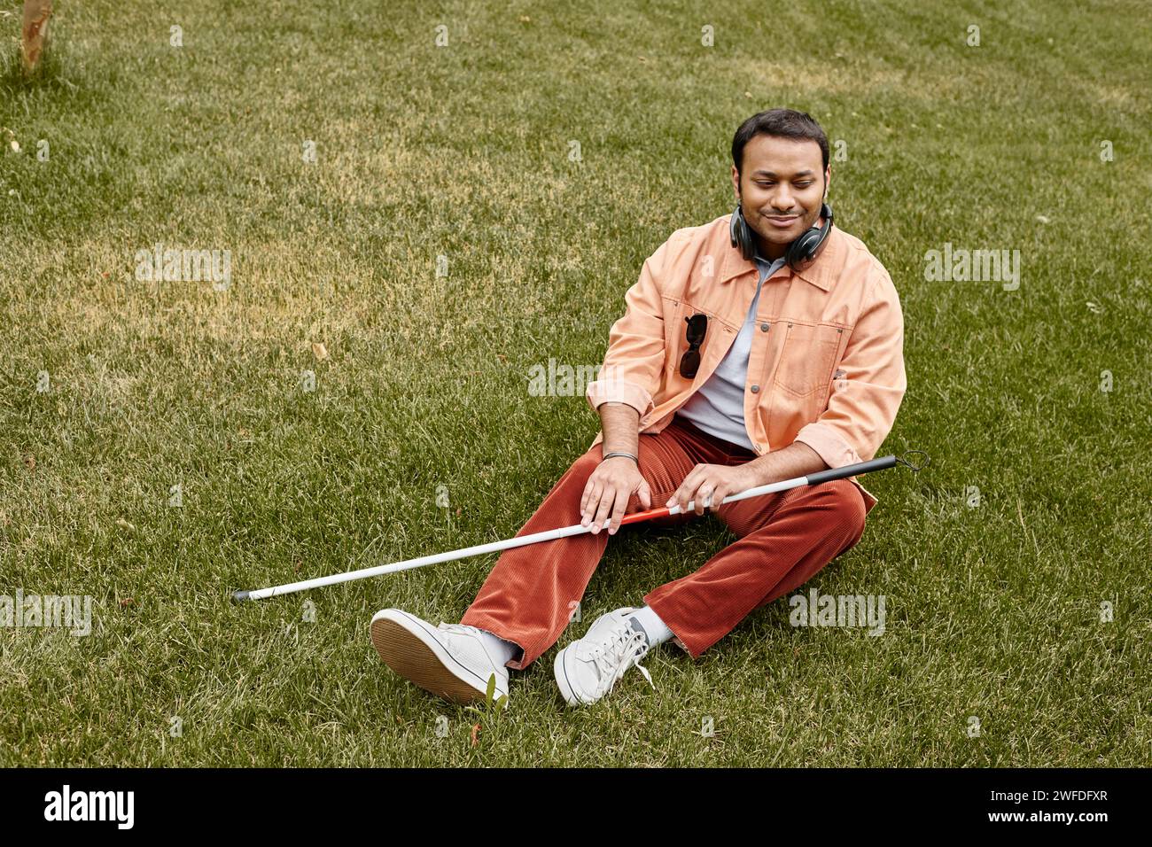 cheerful indian blind man in orange jacket sitting on grass with ...