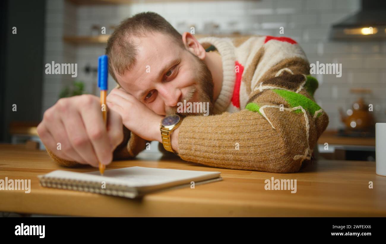 Bored adult man lying on table desk in kitchen drawing in notebook. Man ...