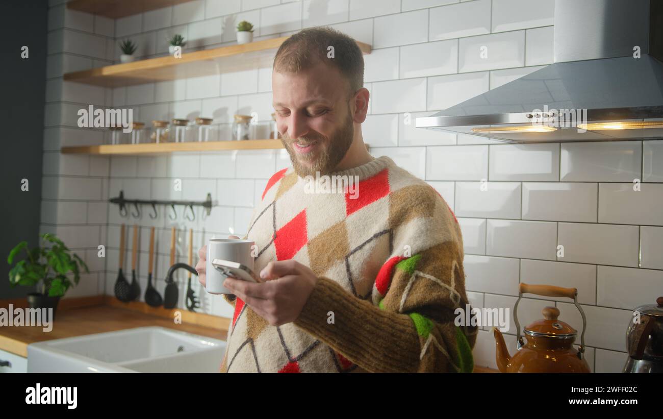 Smiling man standing in modern sunny kitchen having morning coffee scrolling news feed on ...