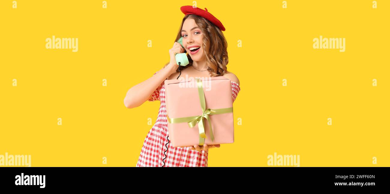 Young woman with gift talking by retro telephone on yellow background ...