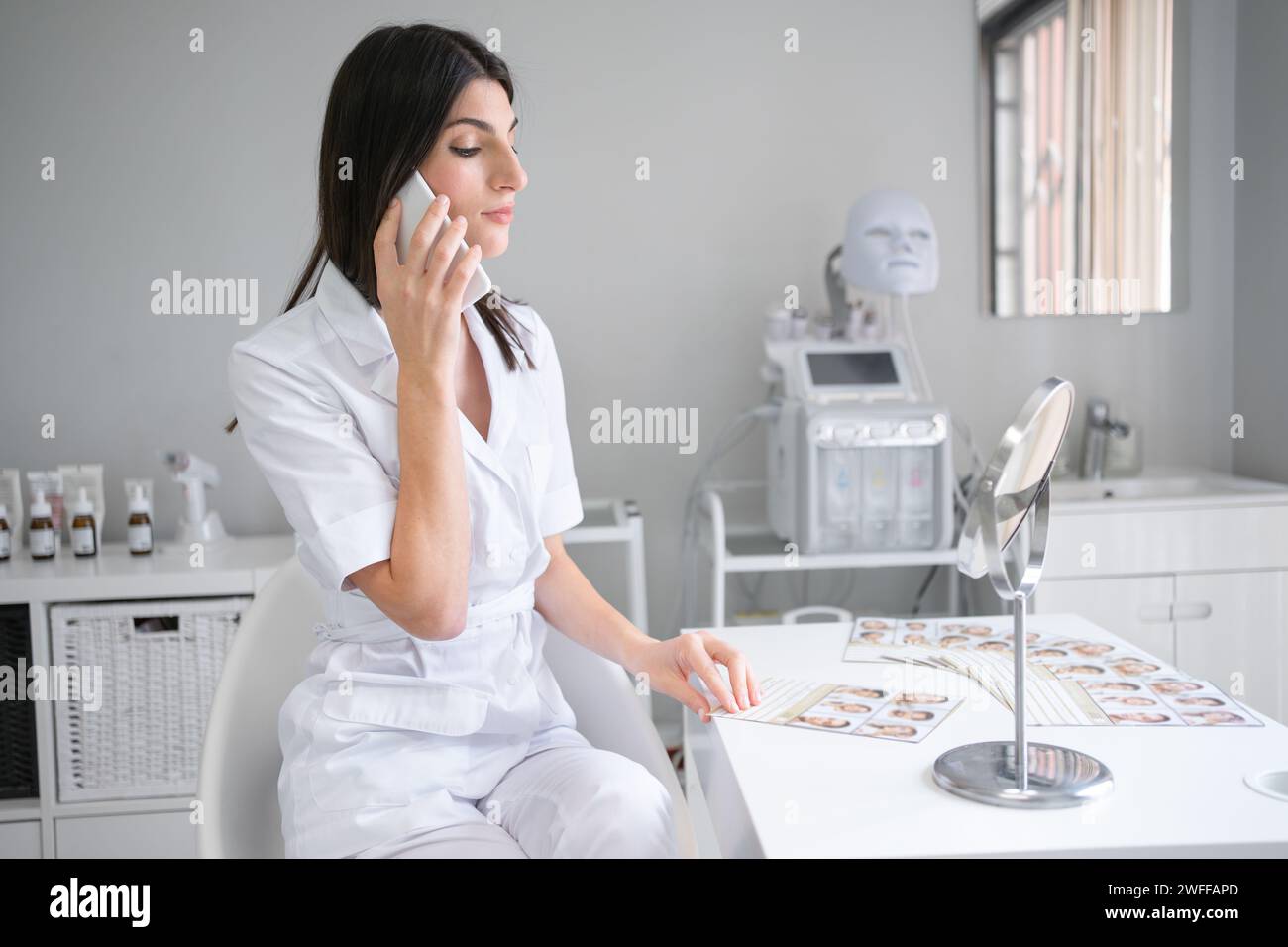 Young female cosmetologist in white uniform talking on smartphone at ...