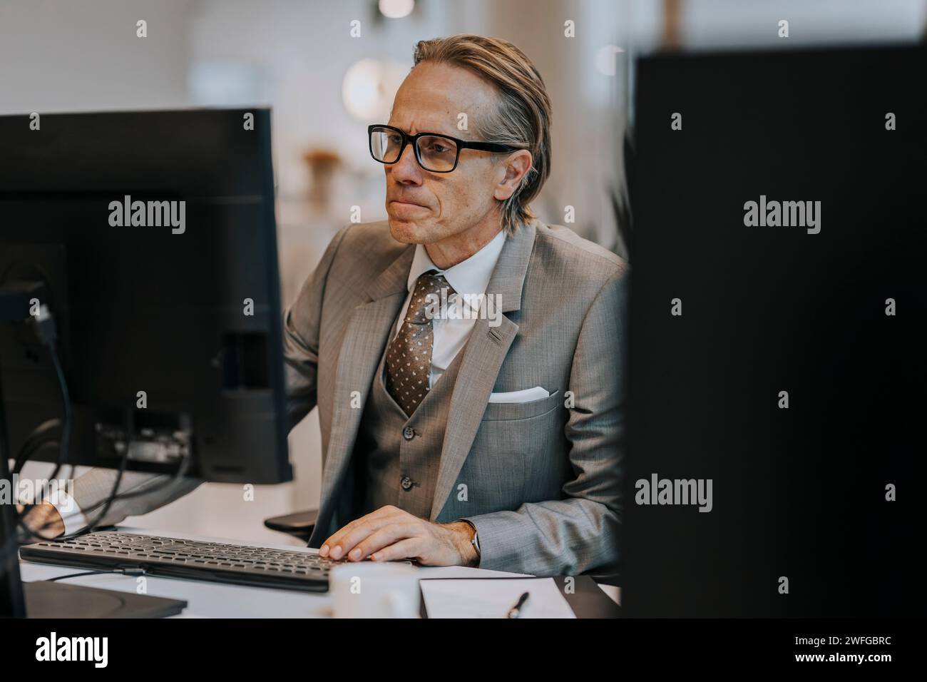Mature businessman wearing eyeglasses using computer at desk in office ...