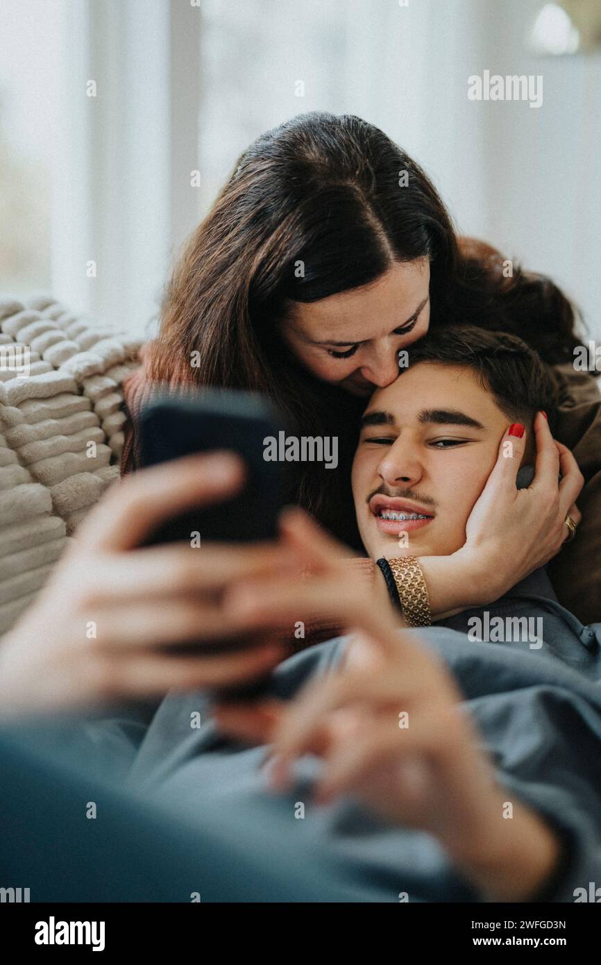 Teenage boy using mobile phone while mother kissing him in living room ...
