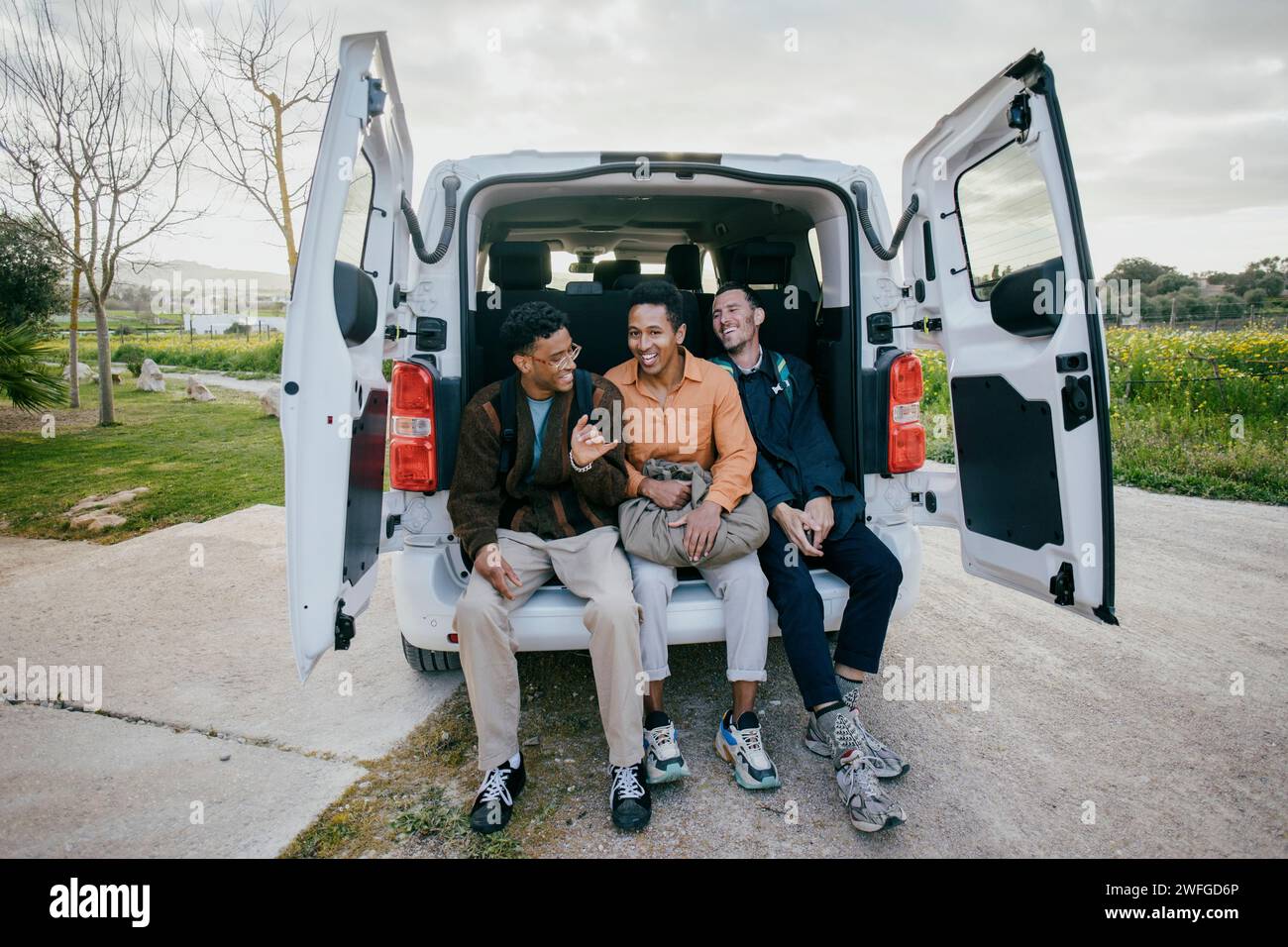 Full length of cheerful male friends sitting in van trunk on roadside ...