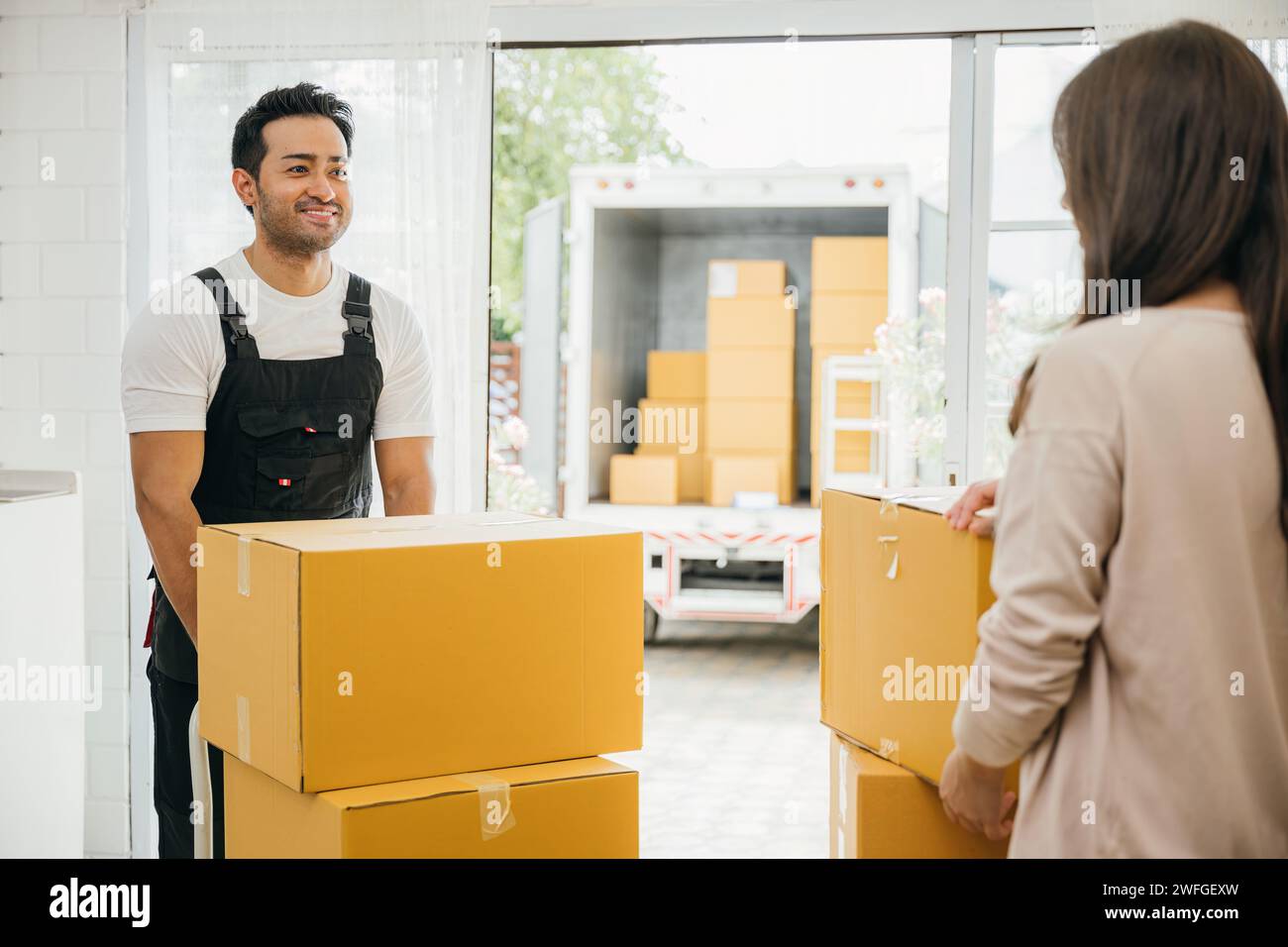 Professional movers assist couple inside home unloading boxes from truck. Service ensures ...