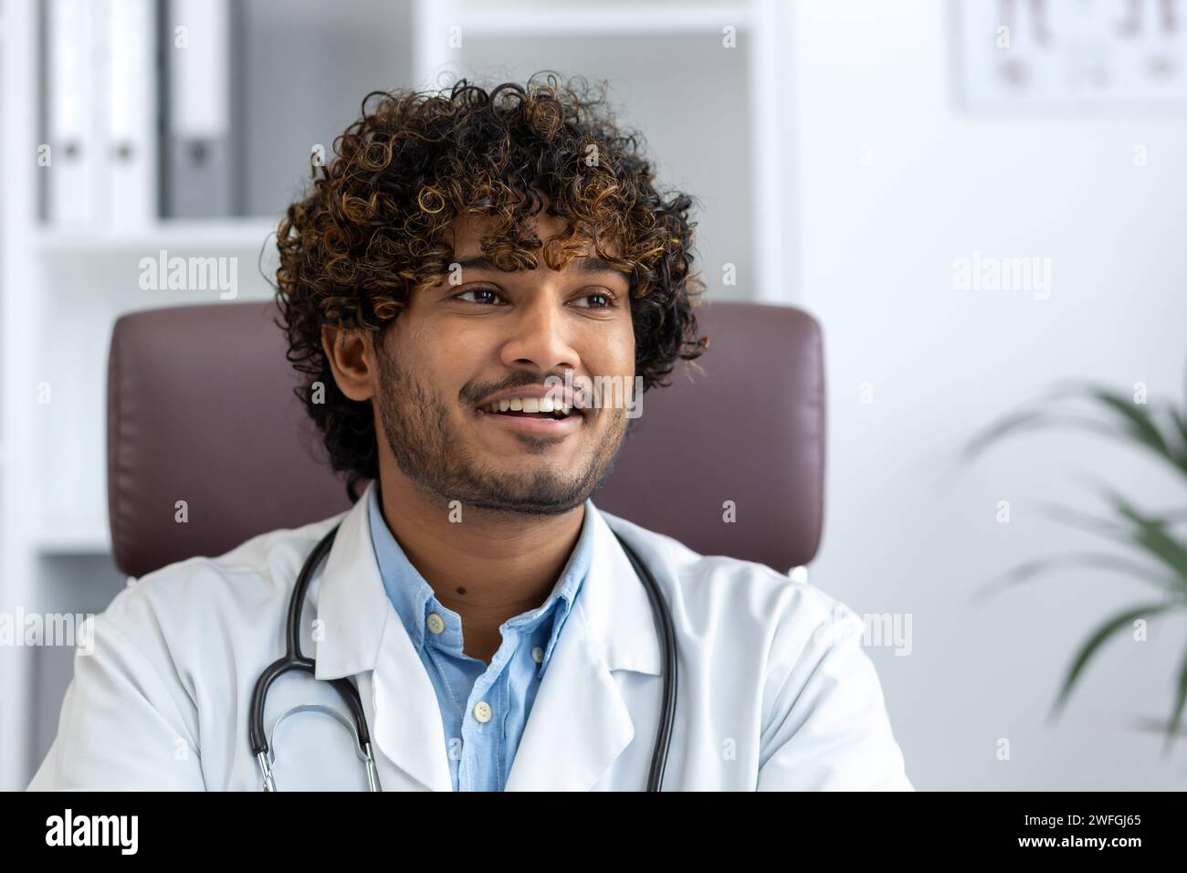 A young male Hindu doctor with curly hair smiling in a medical office ...