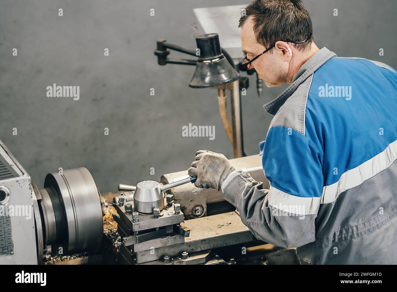 Turner in overalls works at lathe in turning shop at factory. Elderly ...