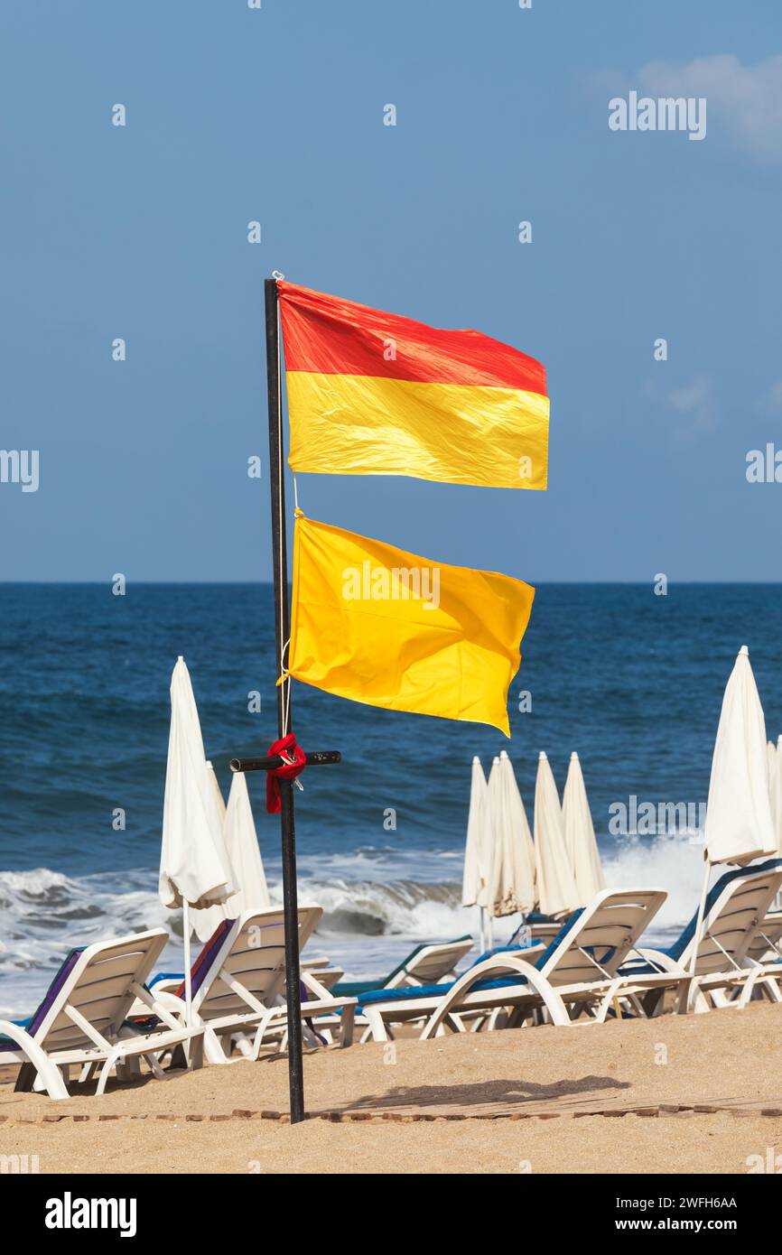 Red and yellow flags flutters on a beach with folded sun loungers and ...