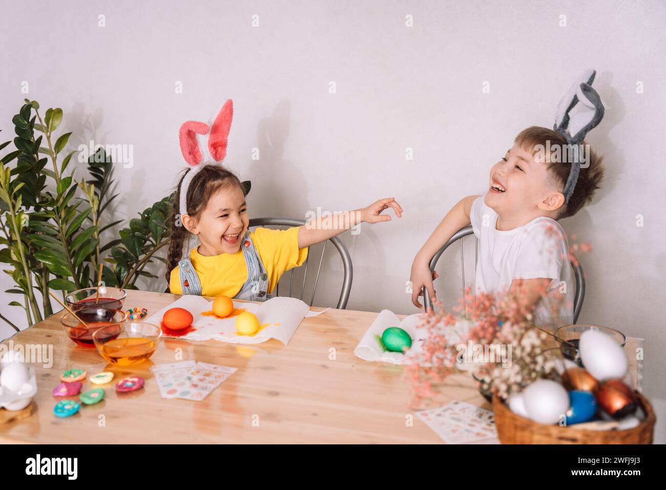Children wearing bunny ears on their heads prepare for Easter by painting eggs in different ...