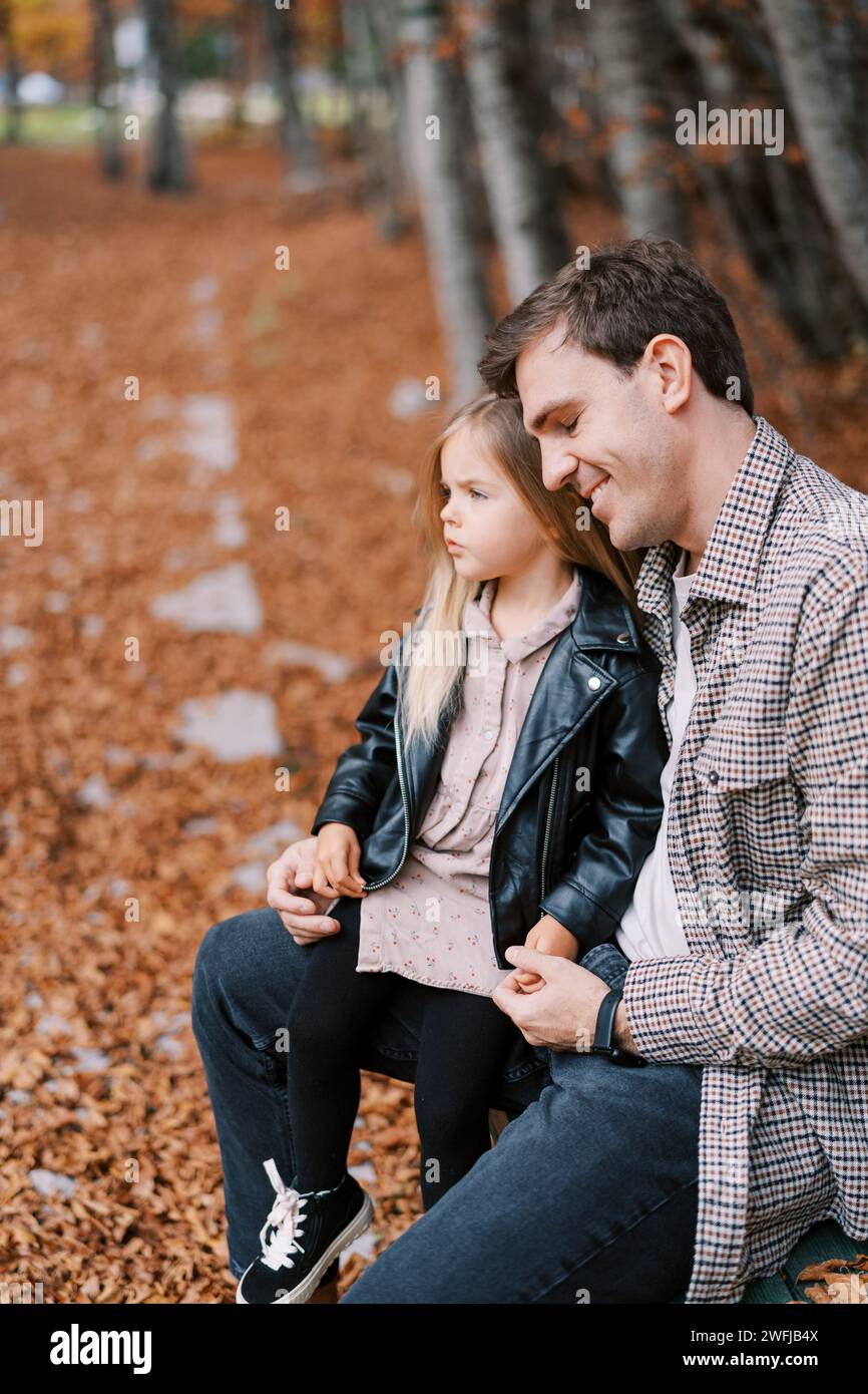 Smiling dad leaned his forehead against the head of a little girl sitting on his lap on a bench ...