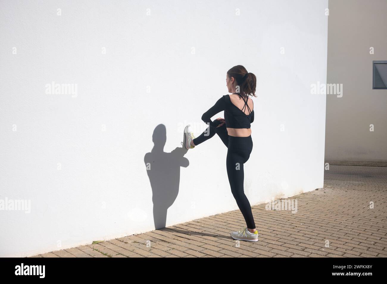 Sporty woman doing a leg stretch exercise against a white wall, warm up ...