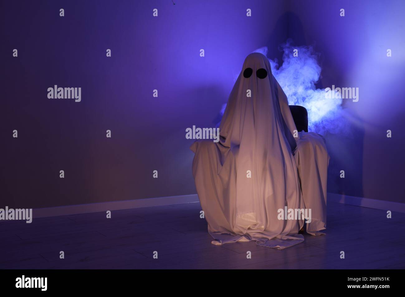 Creepy ghost. Woman covered with sheet sitting in armchair in blue ...