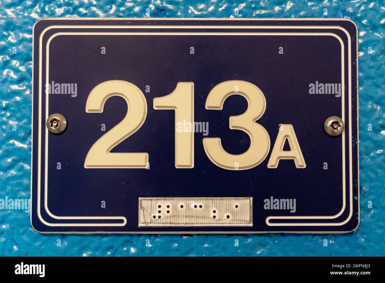Blue 213A sign with white lettering and braille, on a blue block wall ...