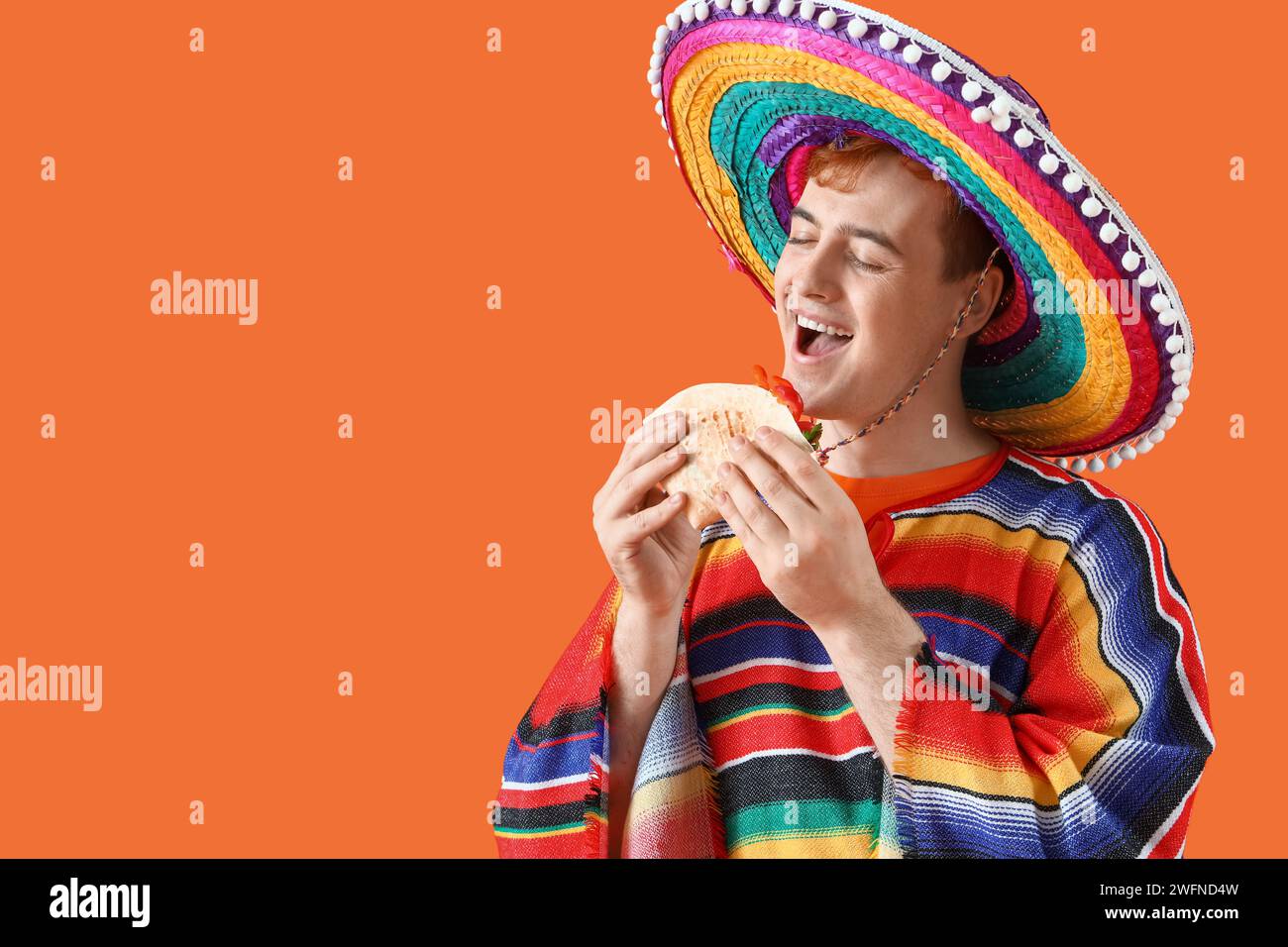 Portrait of young Mexican man in sombrero eating taco on orange ...