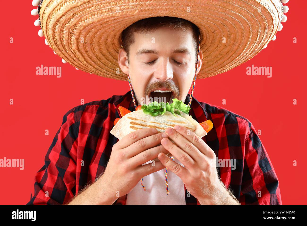 Young Mexican man in sombrero eating taco on red background Stock Photo ...