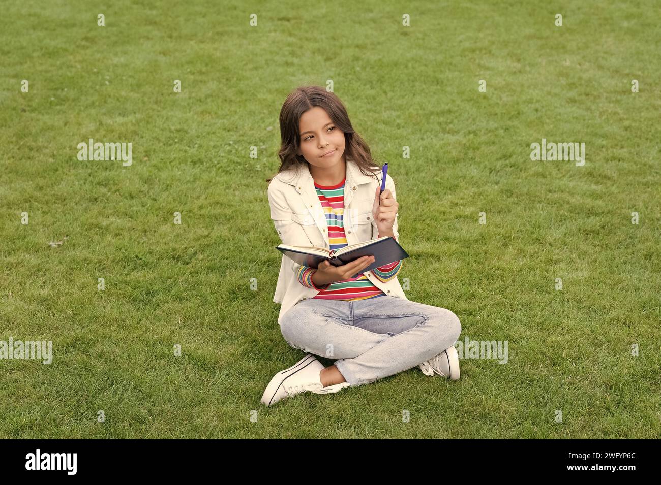 thoughtful teen child making notes in notebook sitting on grass. taking ...