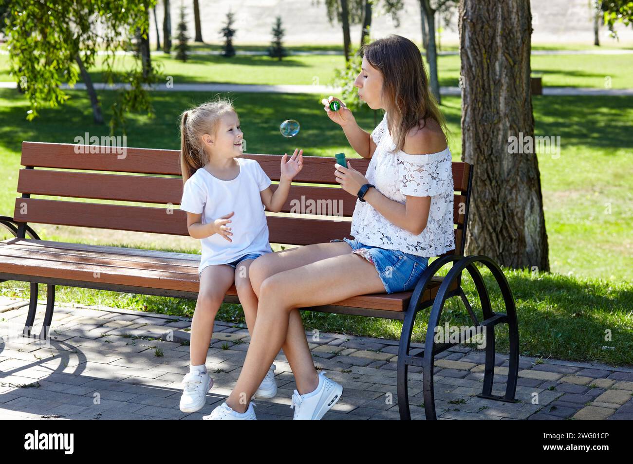 Mother and daughter sitting on a bench in the summer city park and blowing soap bubbles ...