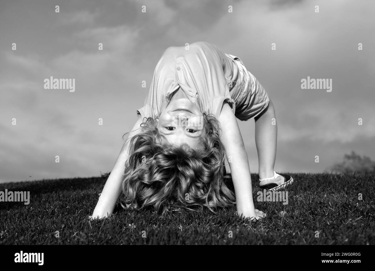 Happy kid boy girl standing upside down on her head on grass in summer day. Funny cute child ...