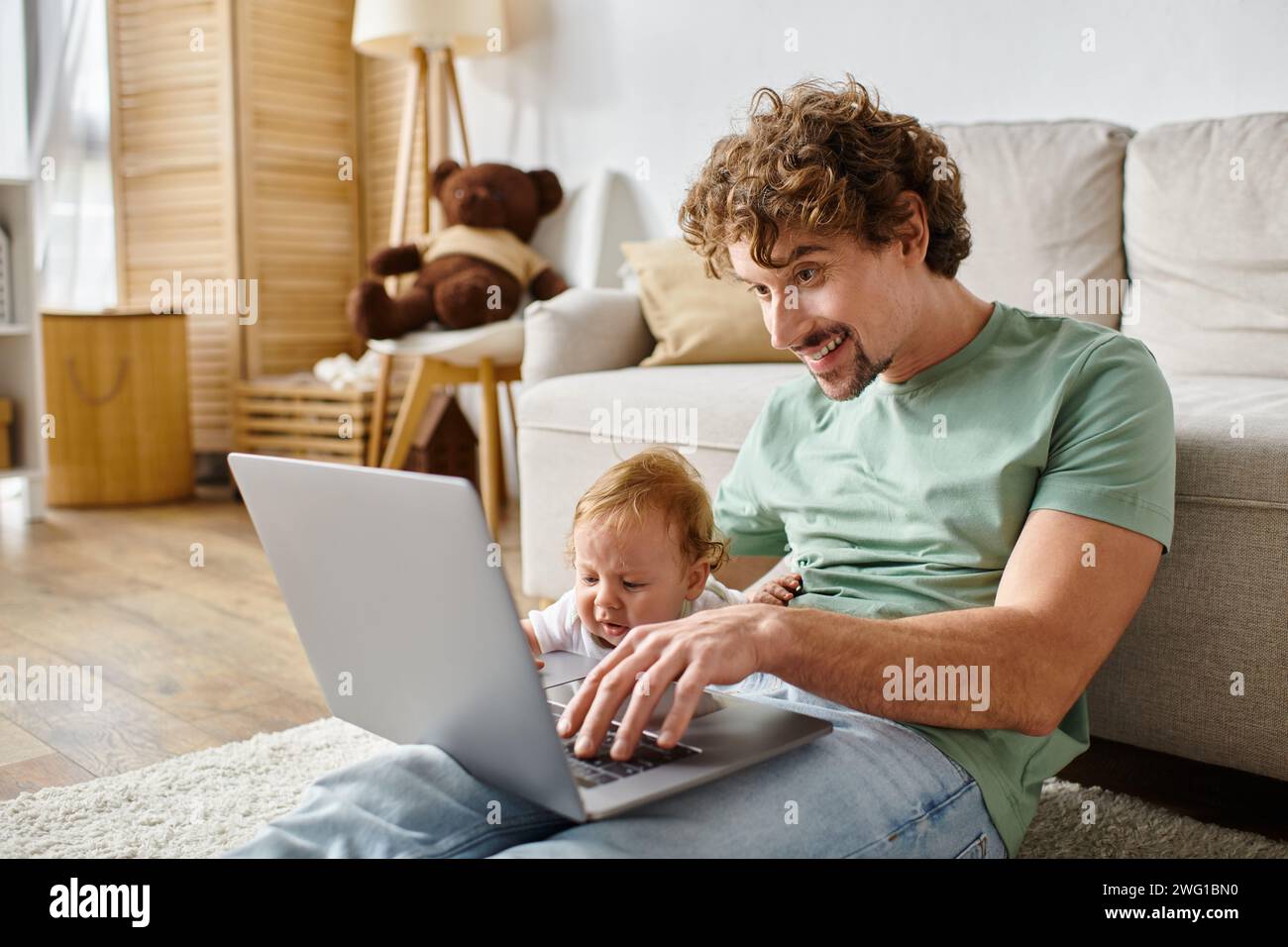 cheerful father using laptop near infant son in living room, balancing ...