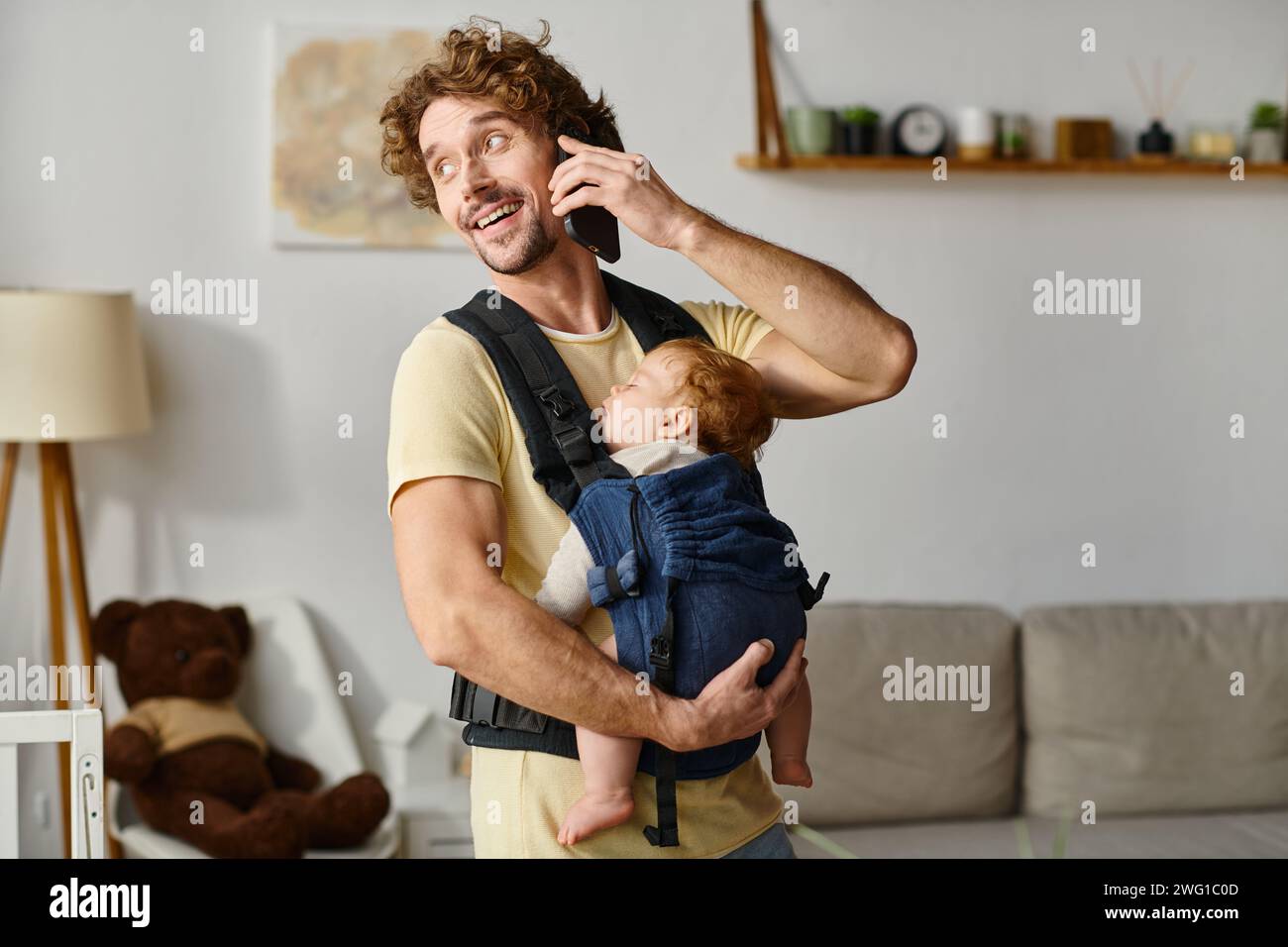 cheerful man talking on smartphone with baby sleeping in carrier ...
