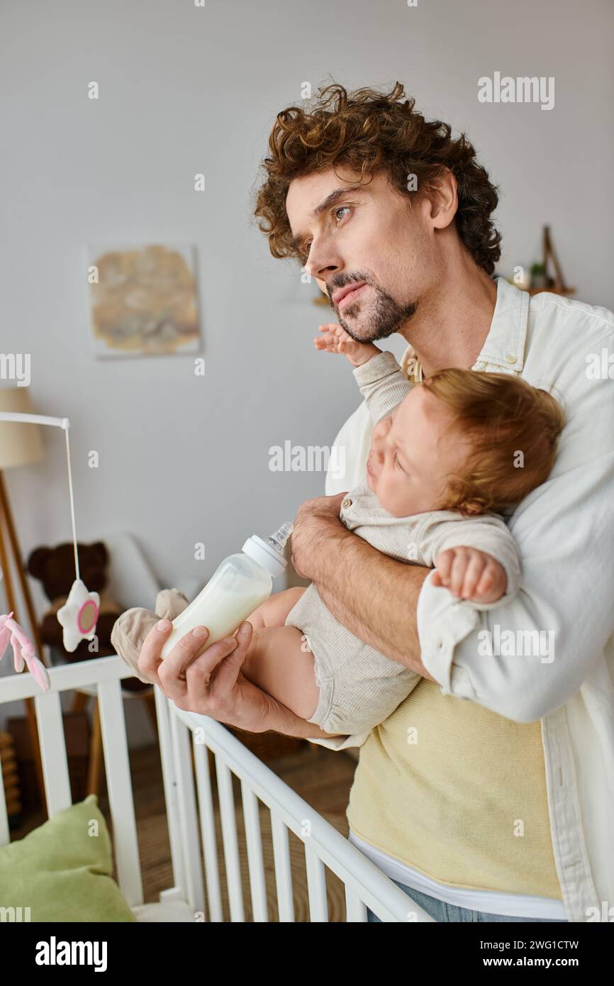 curly-haired father feeding his infant son from baby bottle near crib ...