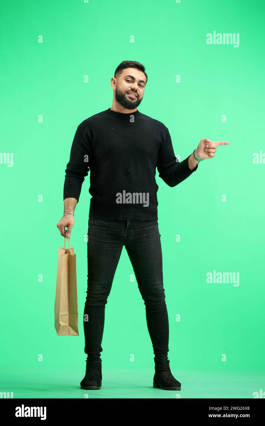 A man, full-length, on a green background, with bags, points to the ...