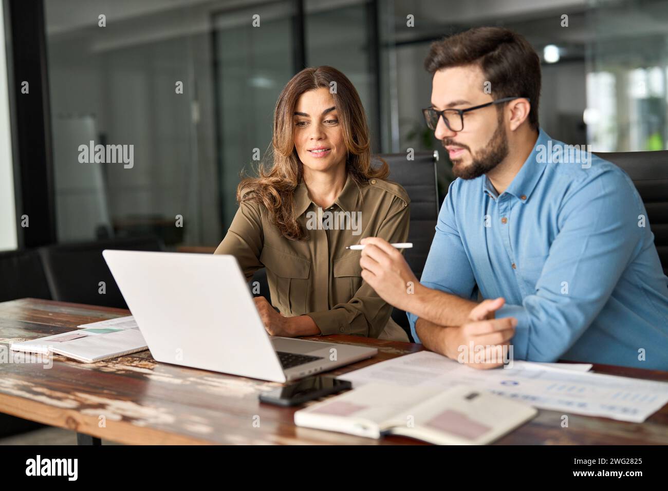 Two busy colleagues working together talking using laptop in office ...