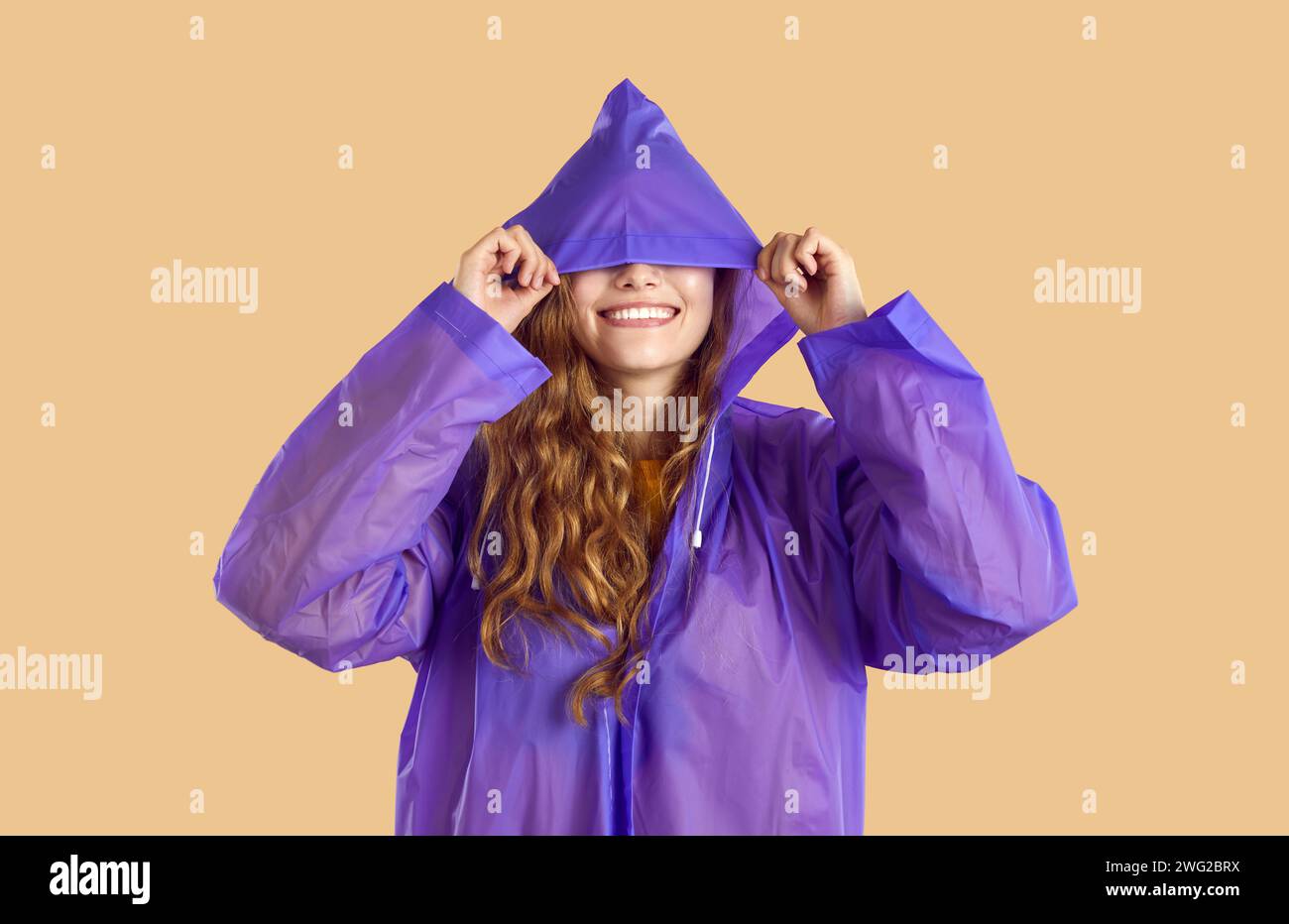 Funny young woman hiding from rain and having fun pulling raincoat hood ...