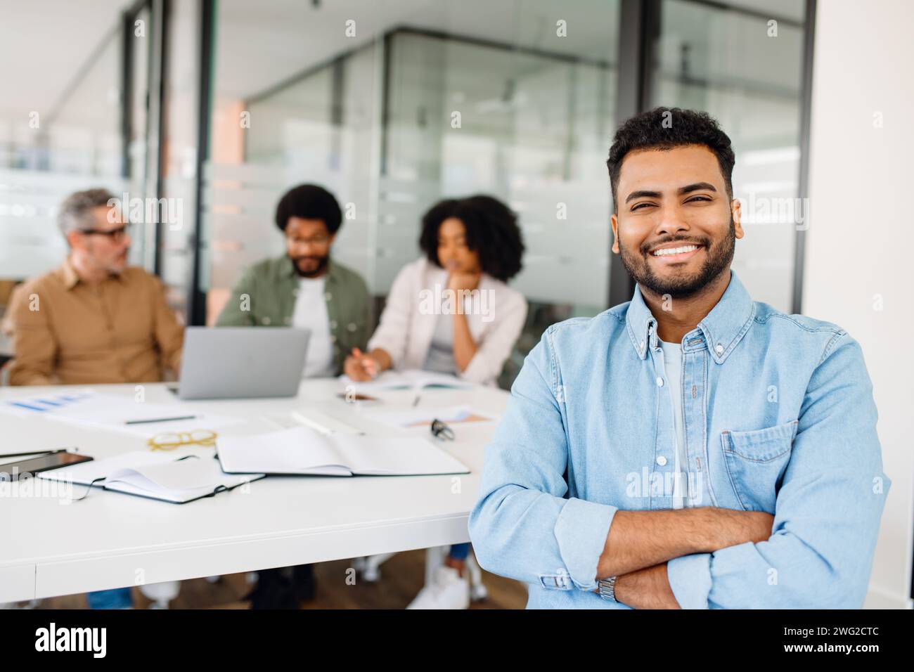 Cheerful Indian professional in a denim shirt stands with arms crossed ...