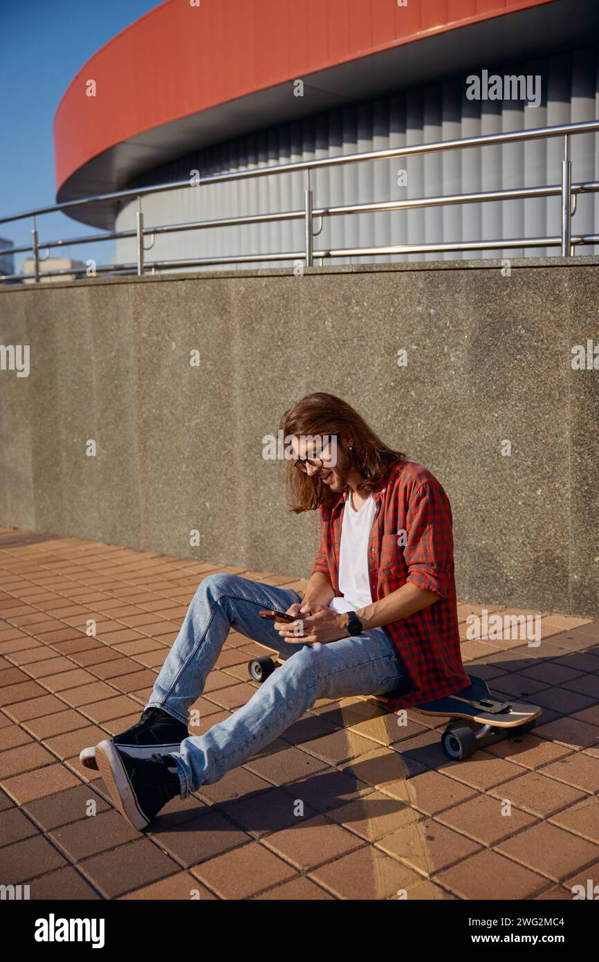 Hipster man with longboard having rest while sitting on steps and using ...