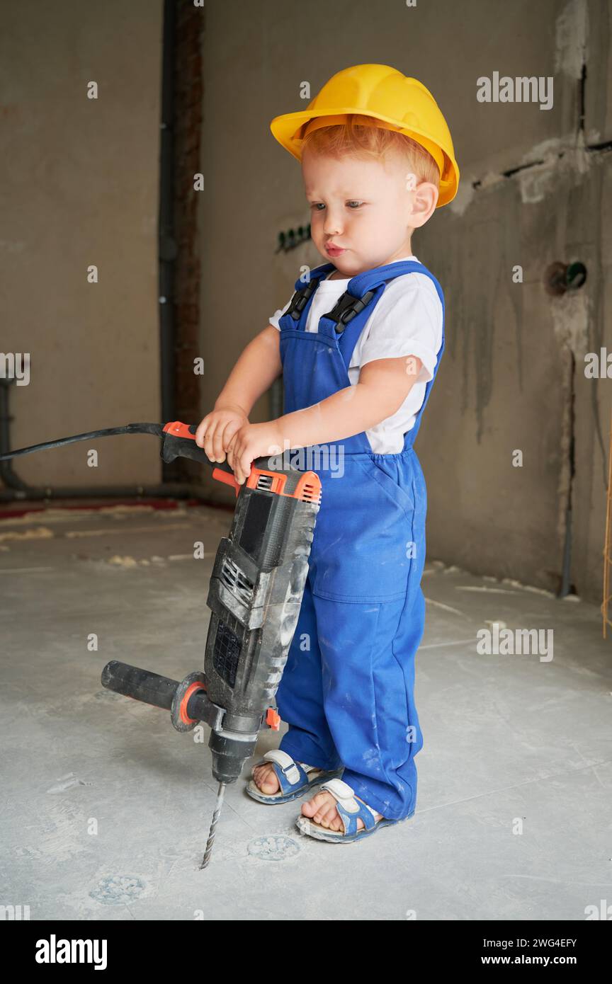 Cute baby boy with electric hammer drill standing in empty room under ...