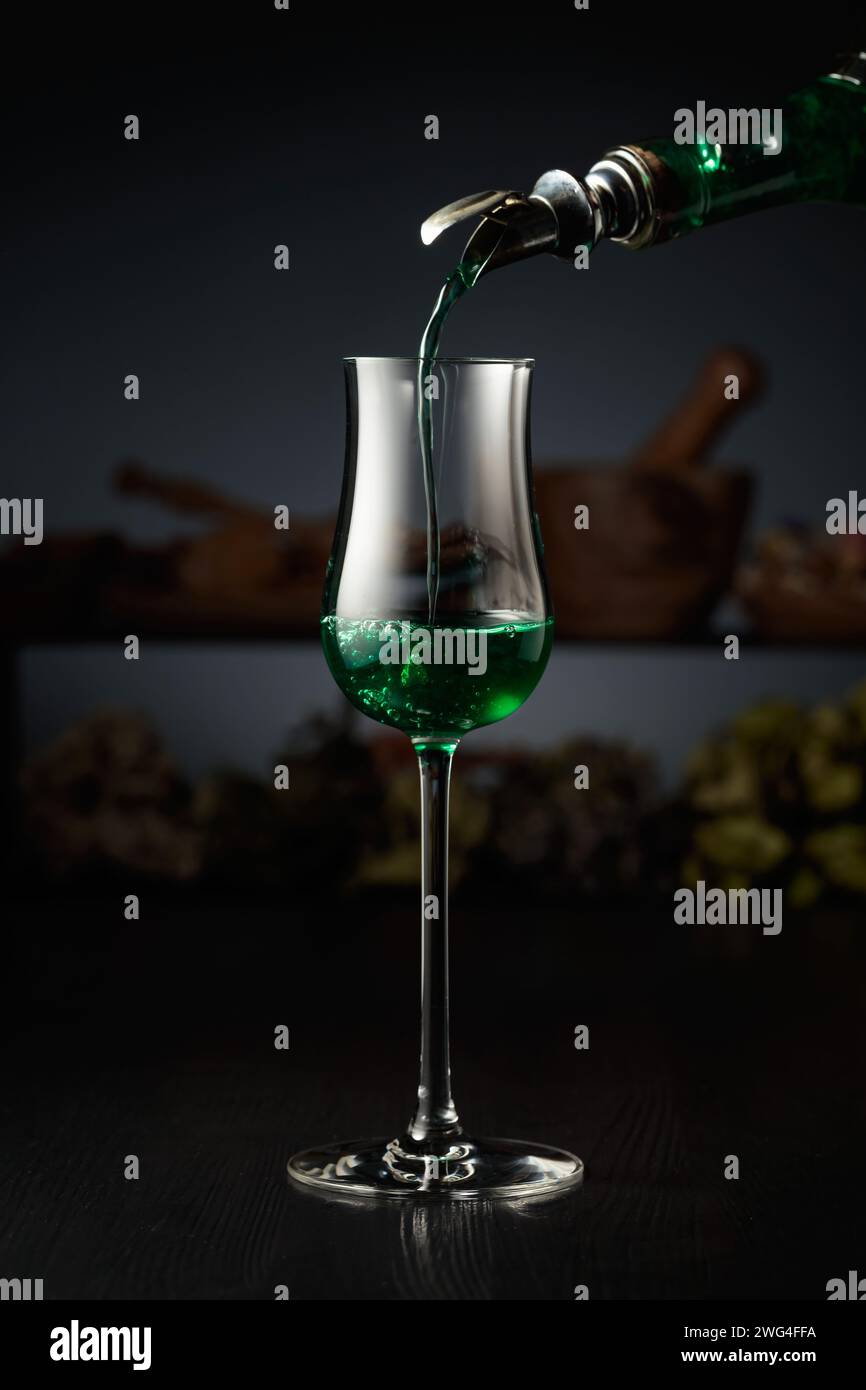 Green herbal liquor is poured from a vintage bottle into a glass. In ...