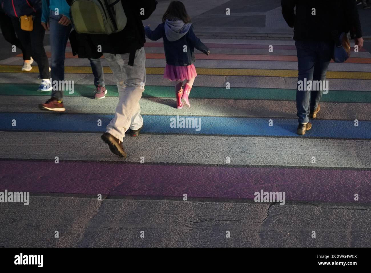 Some people and a child cross a rainbow zebra crossing in the dark ...