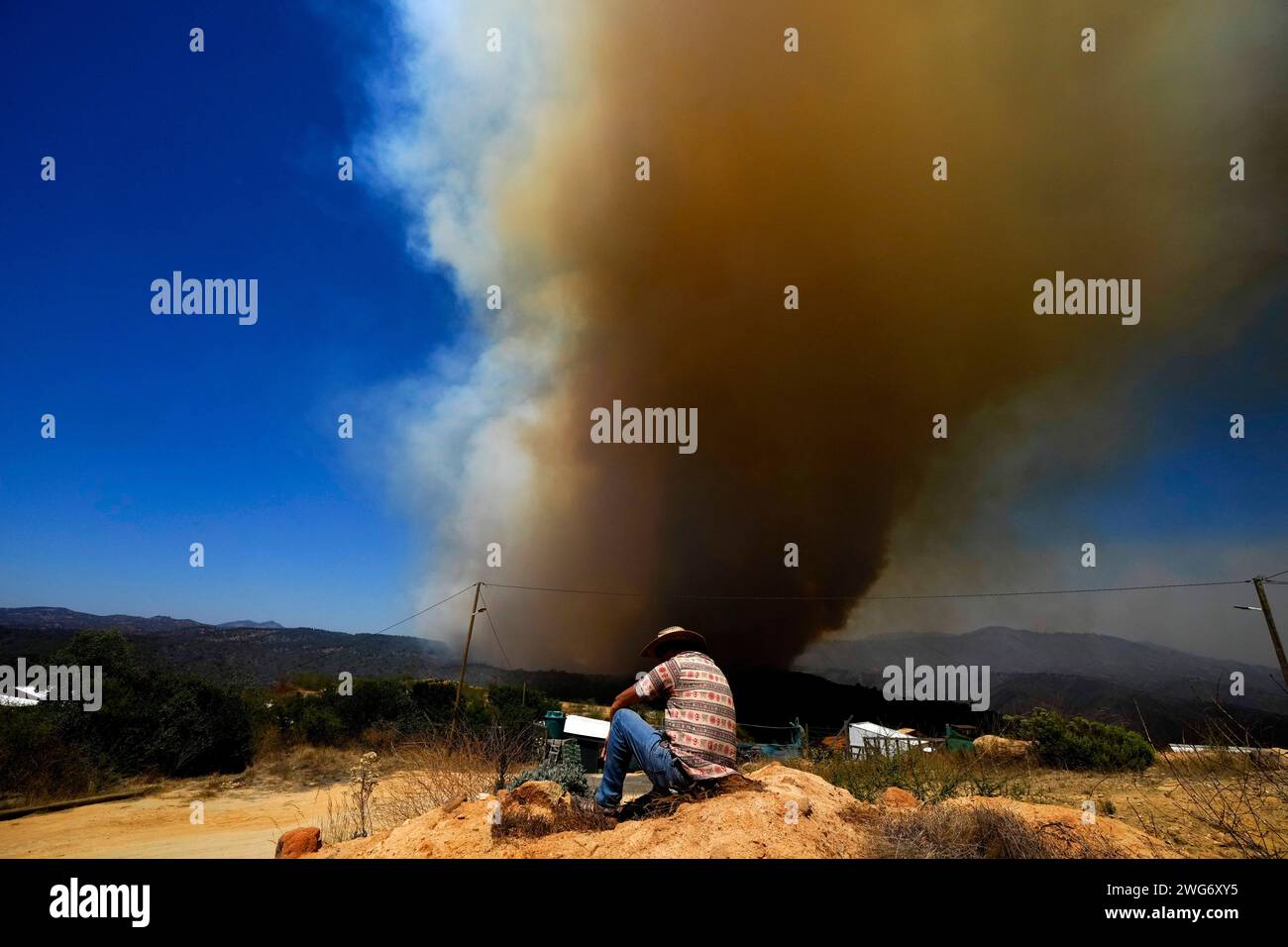 A resident sits on a mound of dirt as a plume of smoke from forest ...
