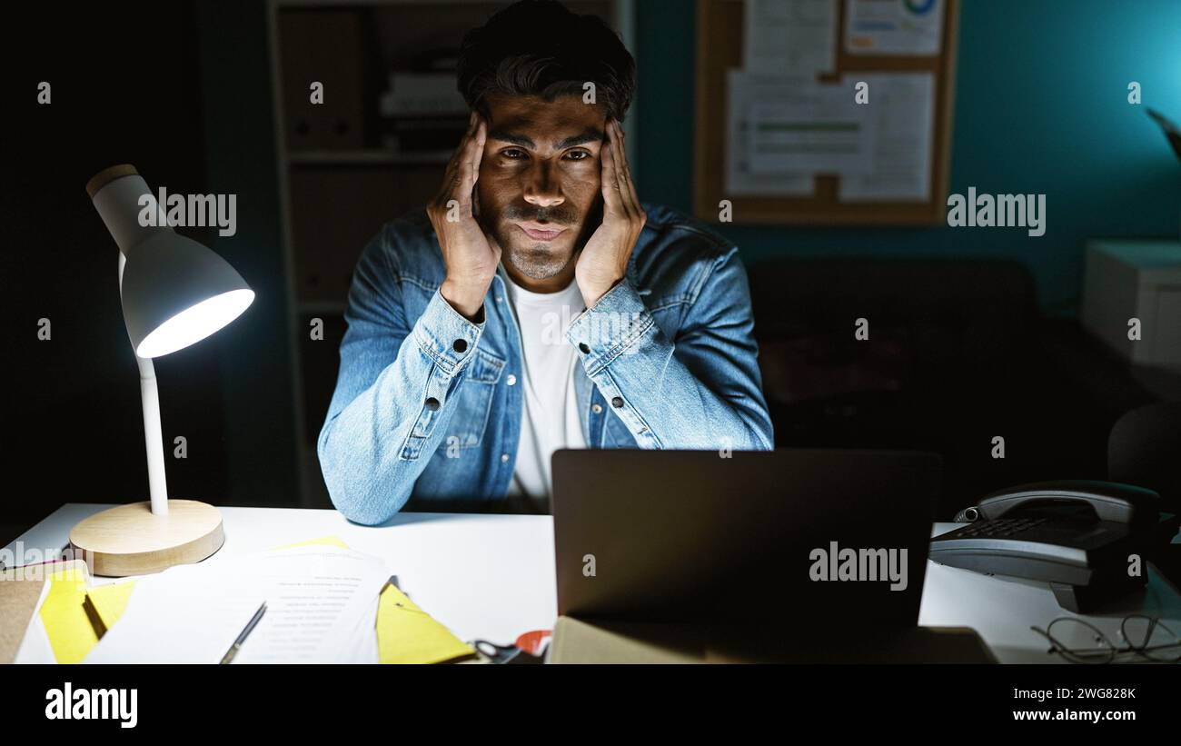 Stressed hispanic man with beard in office late at night, hands on head ...