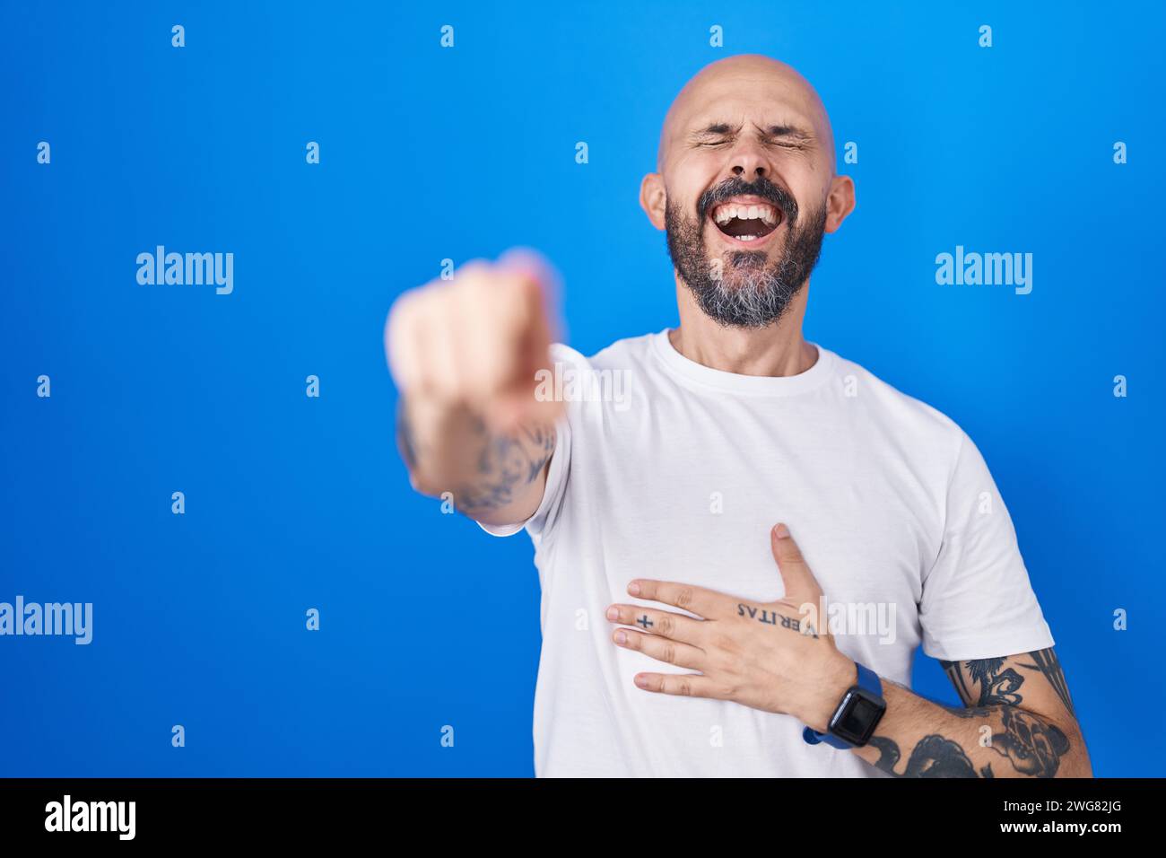 Hispanic man with tattoos standing over blue background laughing at you ...