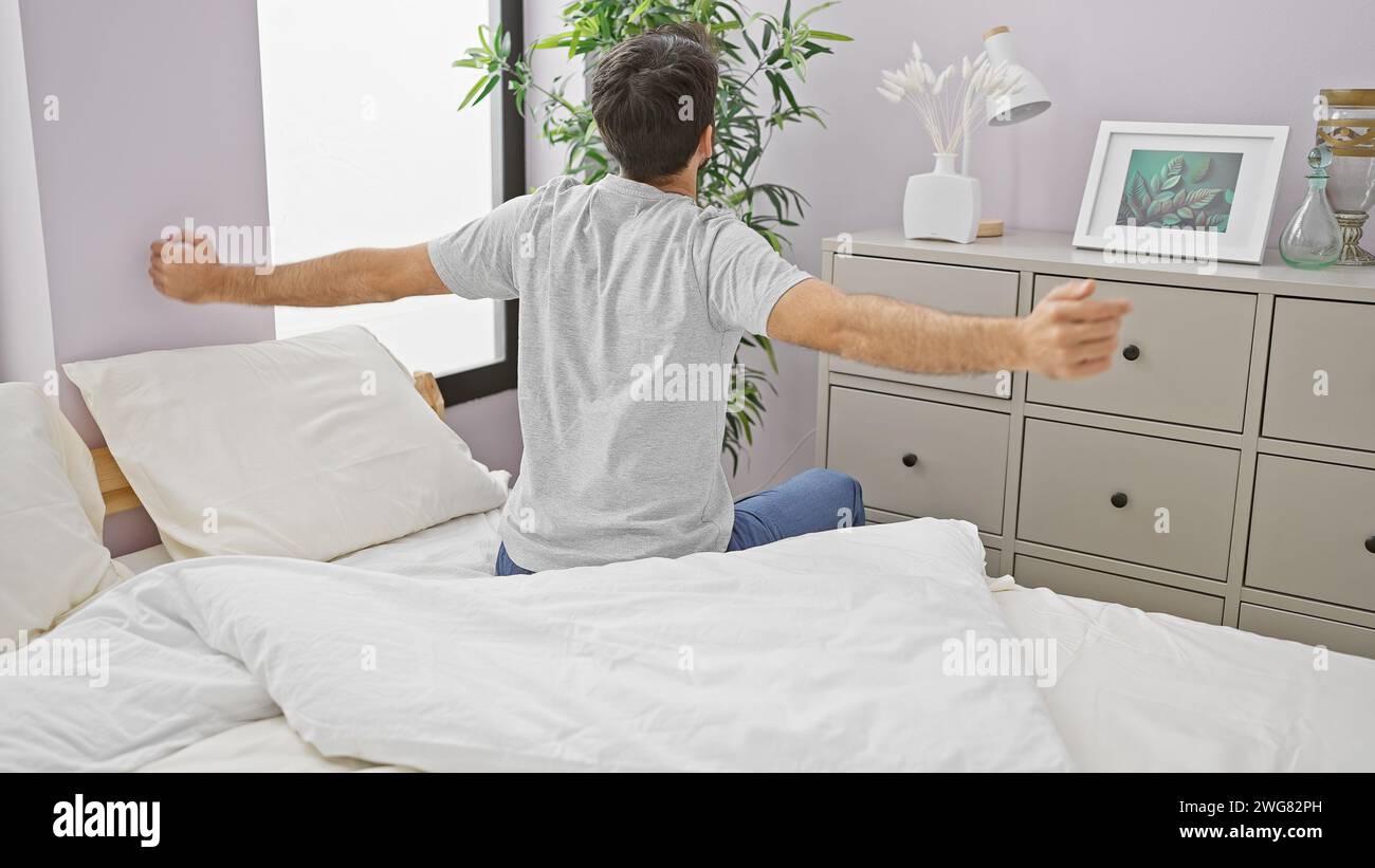 A relaxed young man stretches his arms in a bright bedroom upon waking ...