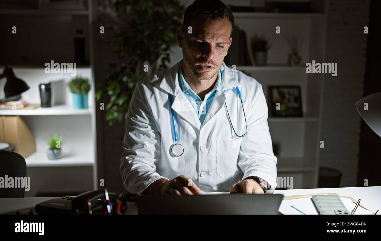 A focused hispanic man in a lab coat works late at night in a clinic ...