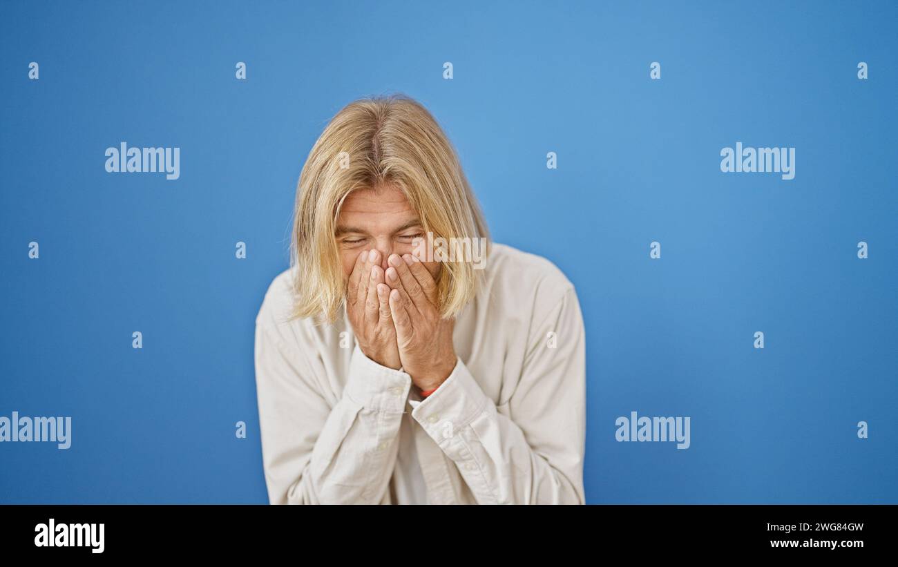 A blond man laughing with hands over his mouth against a blue ...