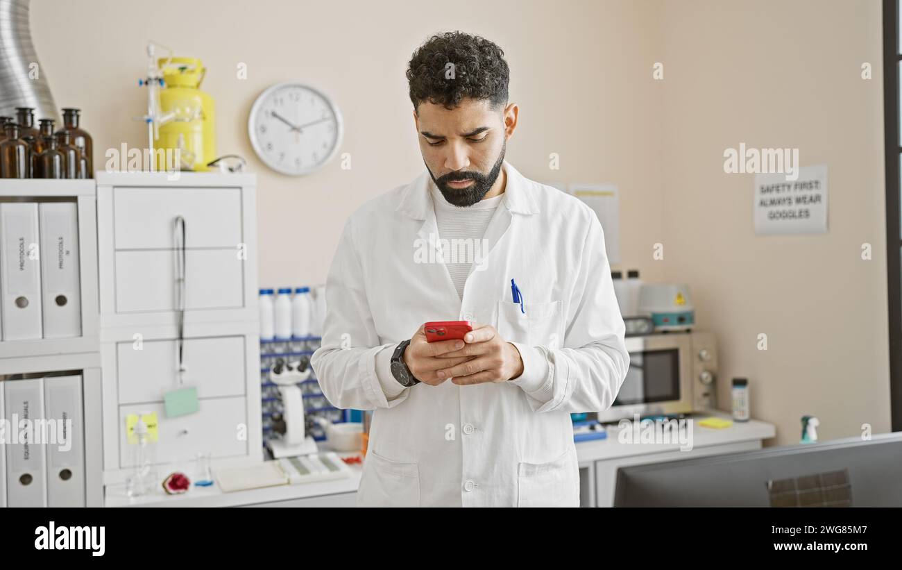Handsome hispanic man in lab coat uses smartphone indoors at a modern ...