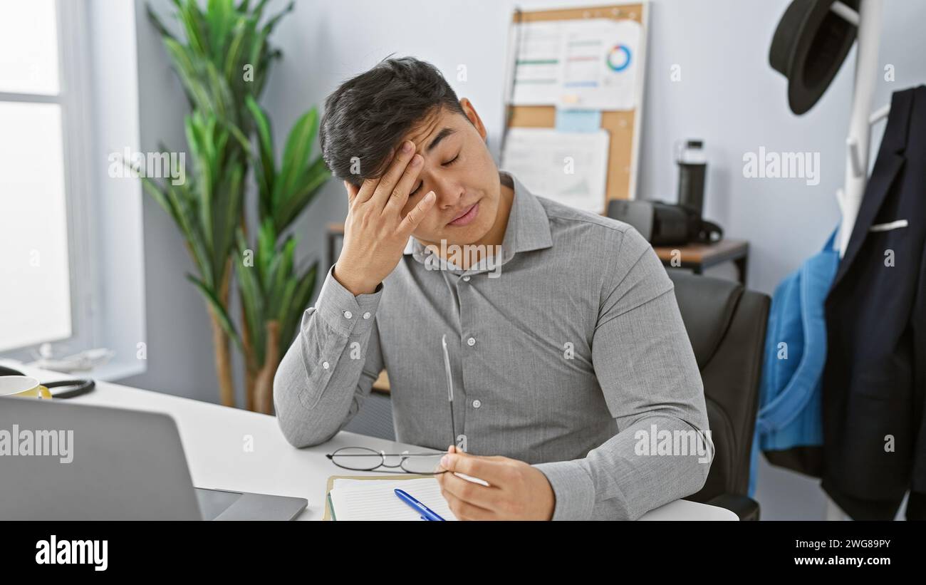 Stressed asian man with hand on forehead sitting at desk in modern ...